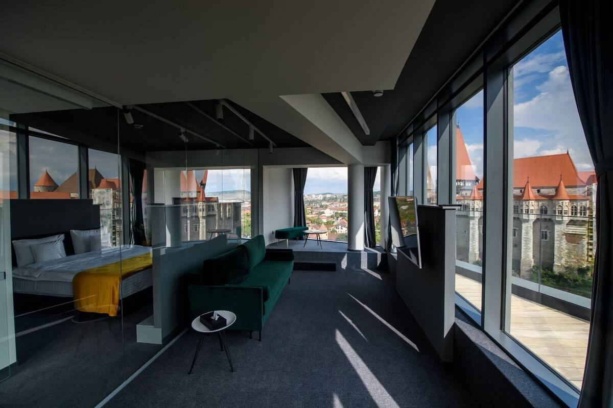 Quarto de hotel moderno com cama, sofá verde e uma pequena mesa. Janelas amplas oferecem vista para um horizonte urbano com edifícios de telhado vermelho.