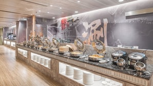 Buffet montado com diversos pontos de comida, incluindo caldeirões quentes e um cozedor a vapor, em um ambiente moderno de hotel.
