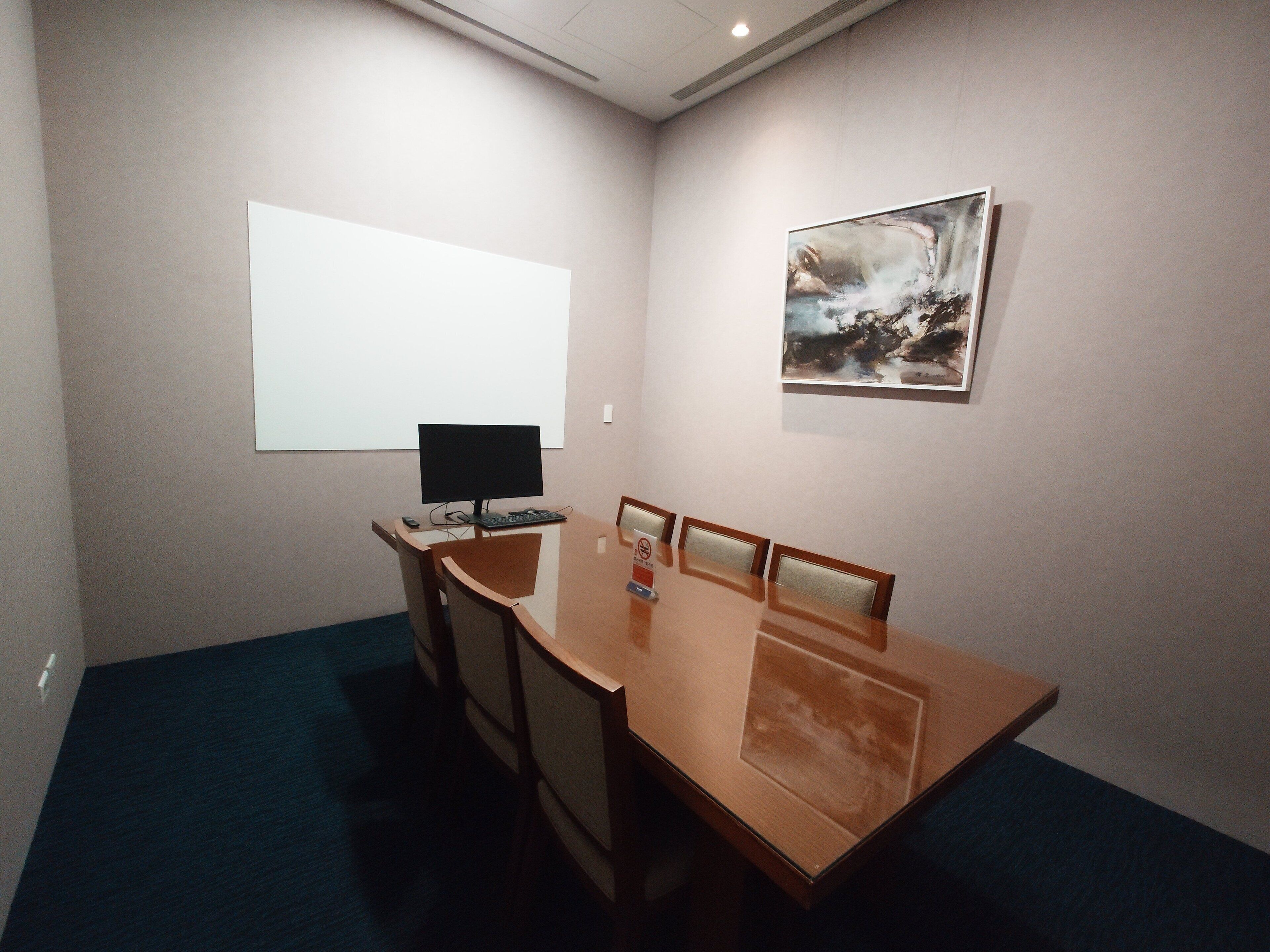 Uma sala de conferências com uma mesa de madeira, quatro cadeiras, uma tela de projetor, um computador e uma obra de arte emoldurada na parede.