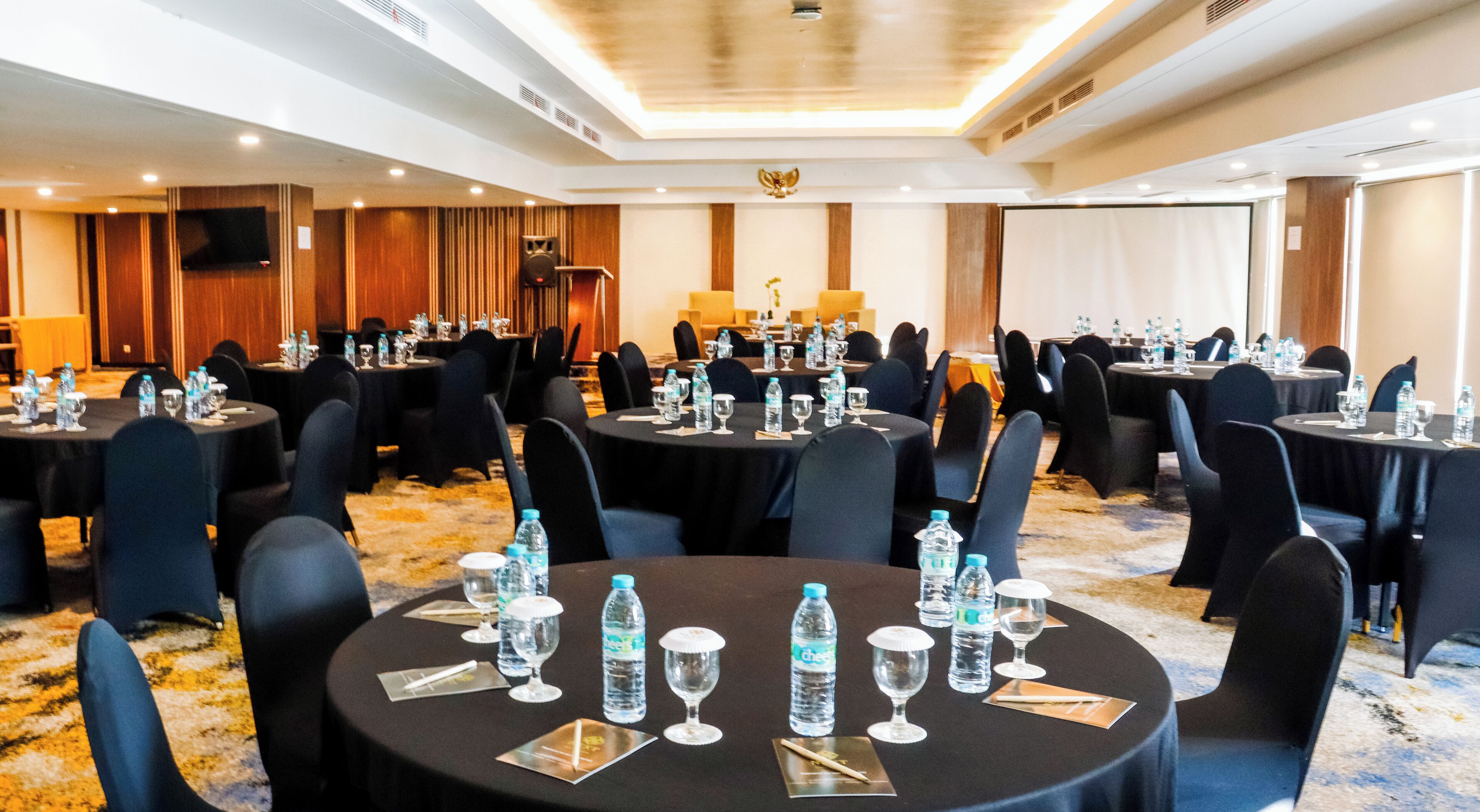 Sala de conferência com mesas redondas, cadeiras pretas e garrafas de água.