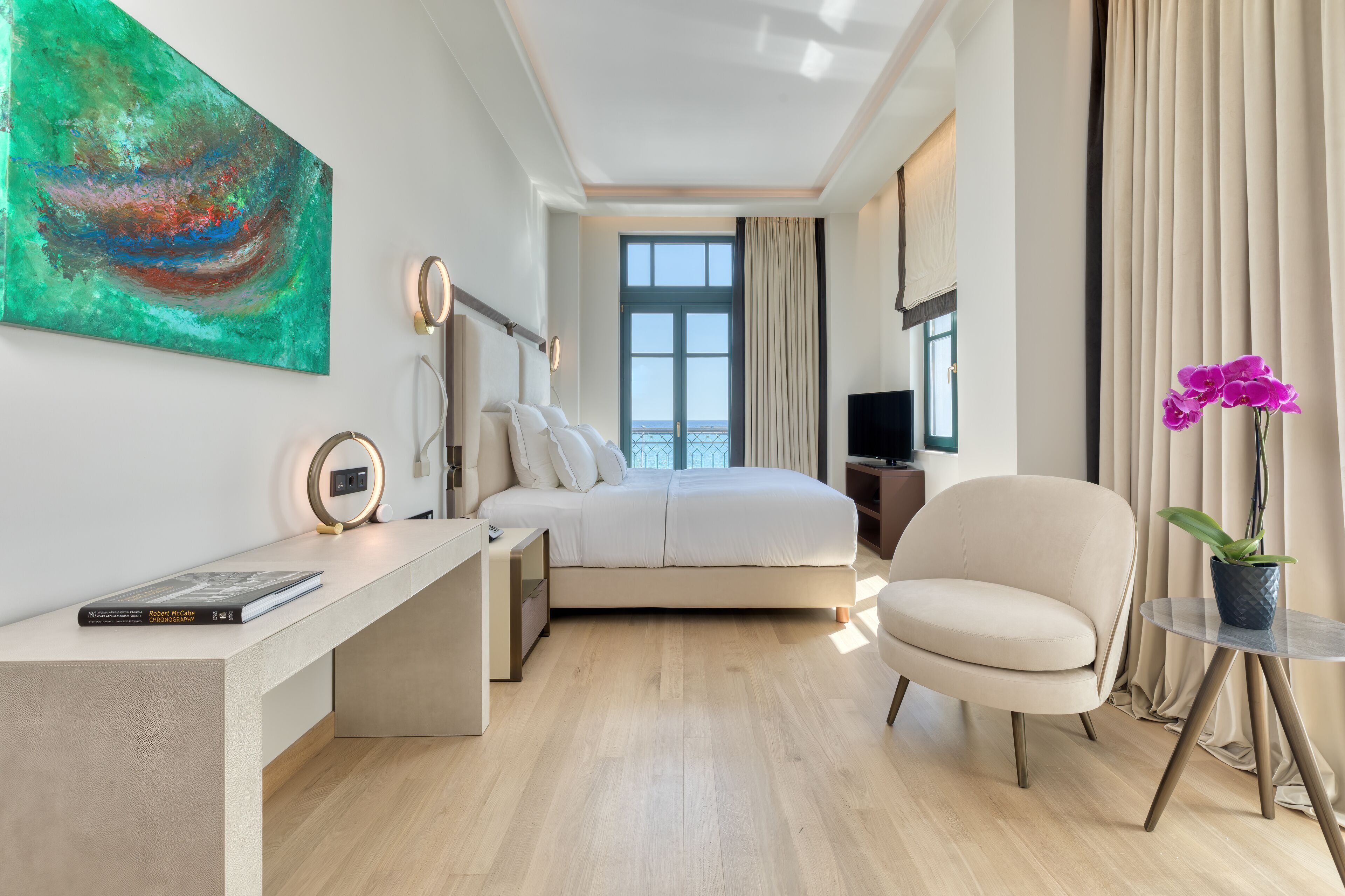 Quarto de hotel moderno com cama, escrivaninha, cadeira, pequena mesa com planta e uma janela grande.
