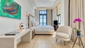 Quarto de hotel moderno com cama, escrivaninha, cadeira, pequena mesa com planta e uma janela grande.