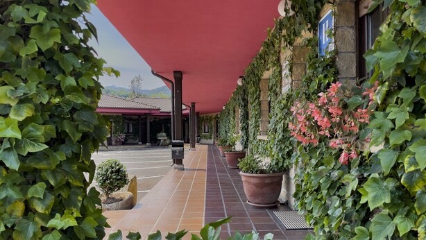 Área externa coberta com vasos de plantas e piso de azulejos, que leva a um edifício com toldo vermelho.