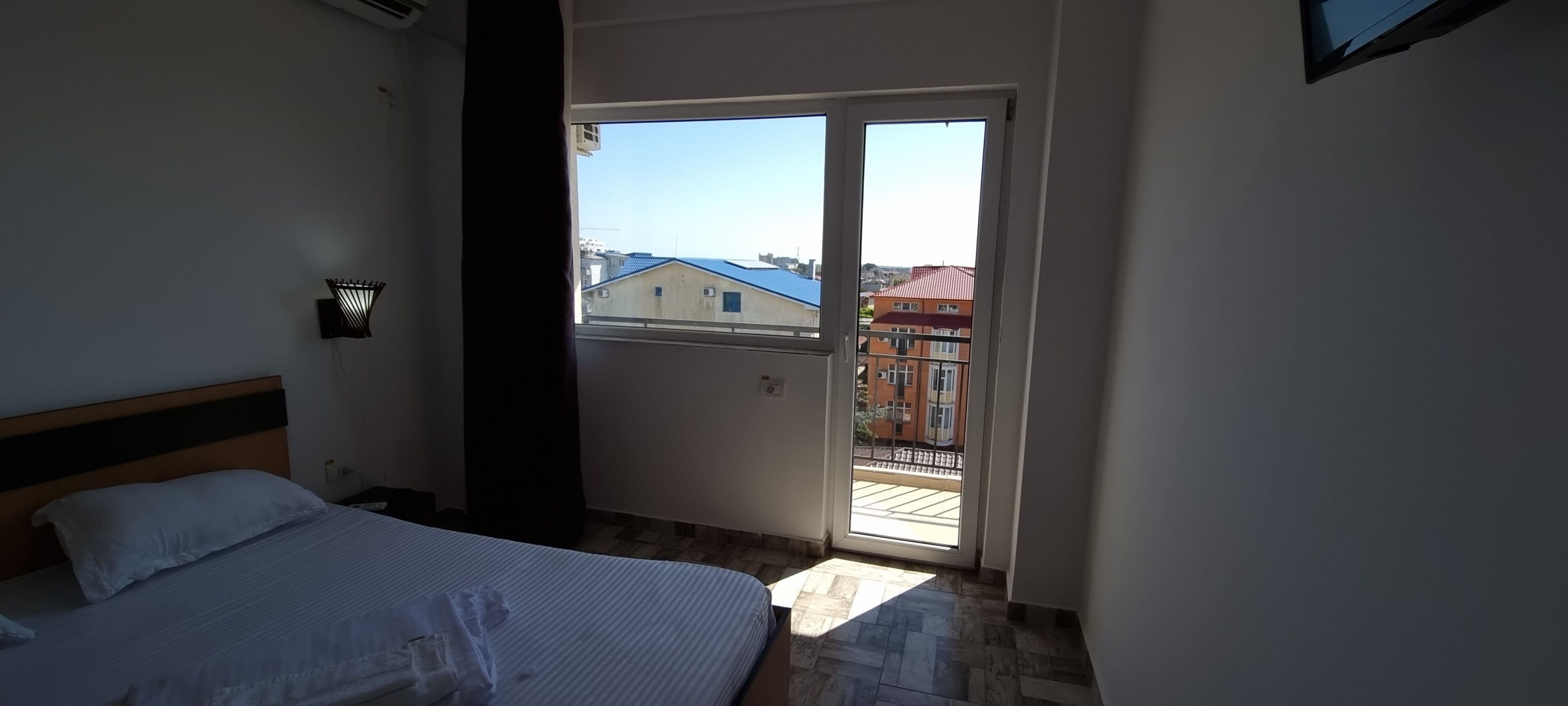 Quarto com cama, mesa de cabeceira, luminária na parede e porta de vidro deslizante para uma varanda externa.