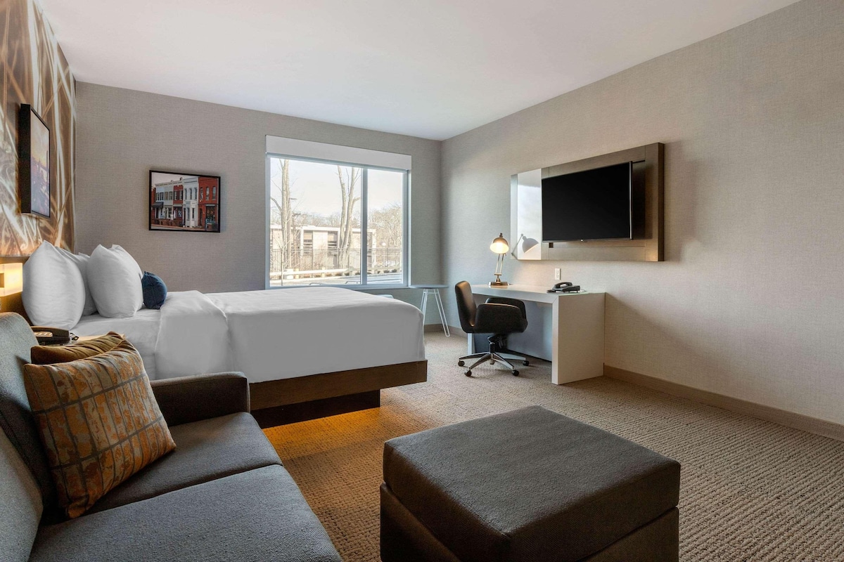 Quarto de hotel com cama, escrivaninha, cadeira, sofá e televisão.