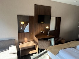 Quarto de hotel com uma cama, escrivaninha, cadeira, televisão na parede e uma luminária.