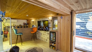 Quarto aconchegante com paredes de madeira e uma grande janela. Há um espaço de bar com banquetas e uma televisão fixada na parede.