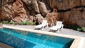 Piscina retangular com padrão de azulejos, rodeada por um deck de pedra e espreguiçadeiras brancas.
