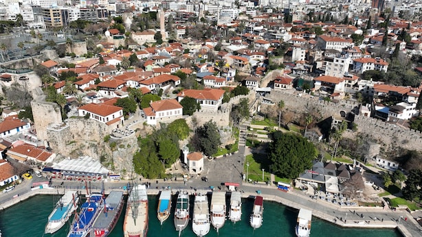 Vista aérea de uma cidade costeira com edifícios históricos, uma marina com barcos e montanhas ao fundo.