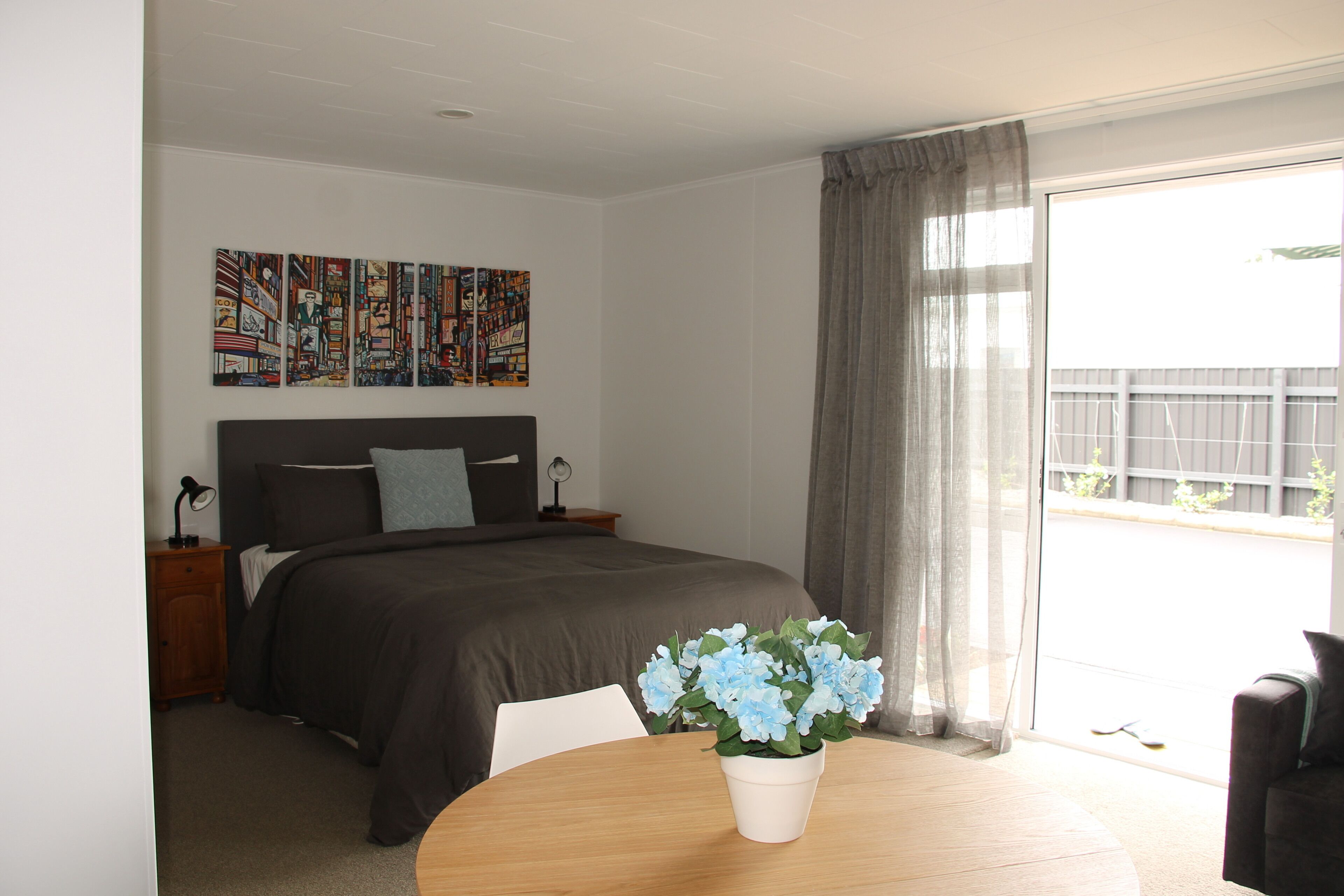 Quarto com cama, mesa de cabeceira, luminária, cadeira, mesa com planta em vaso e uma janela grande com cortinas.