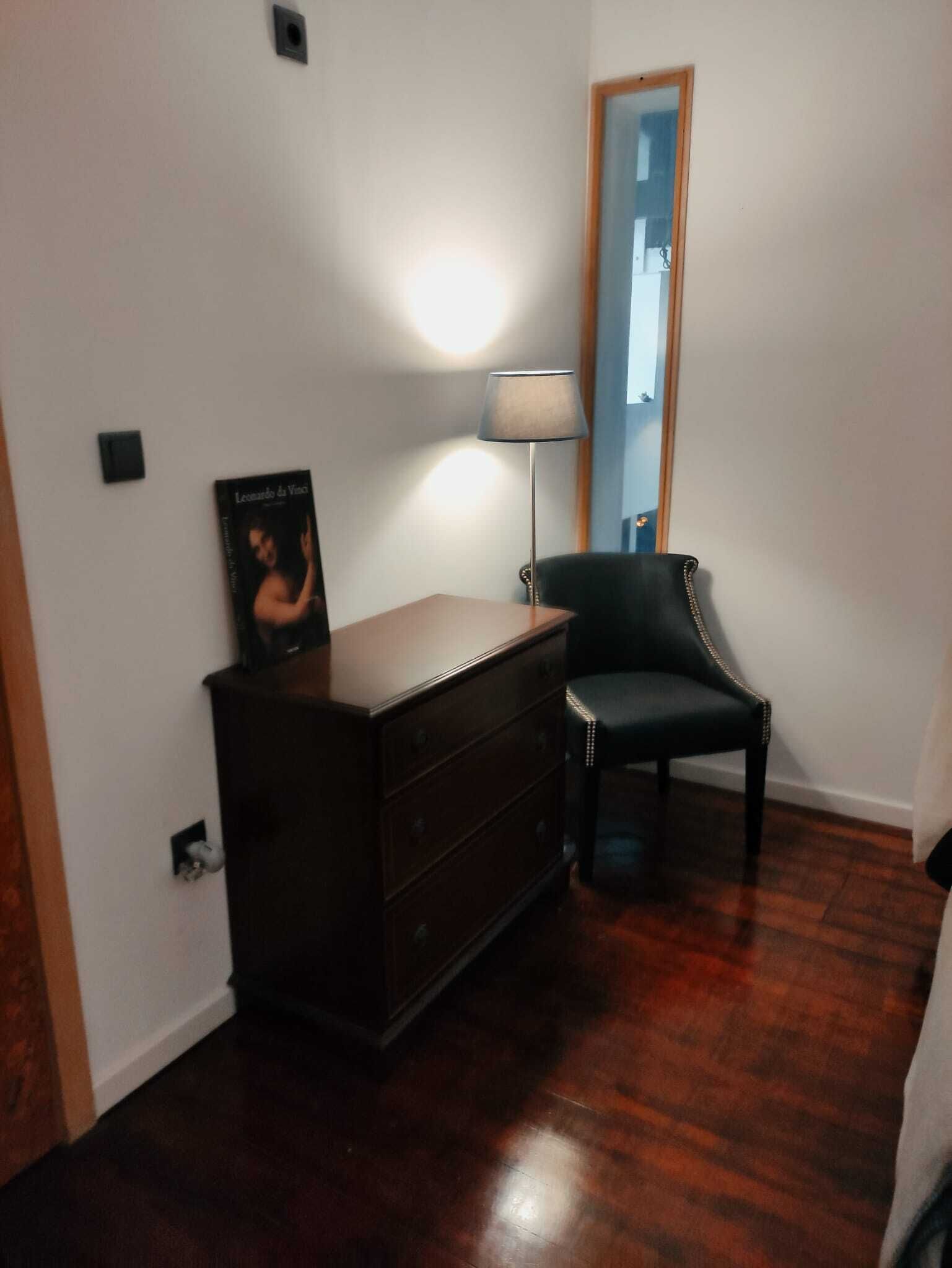 Quarto com cômoda de madeira escura, cadeira preta e luminária de chão.
