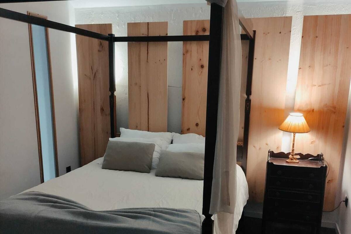 Uma cama com dossel e cortina, mesa de cabeceira com abajur e painéis de madeira nas paredes.