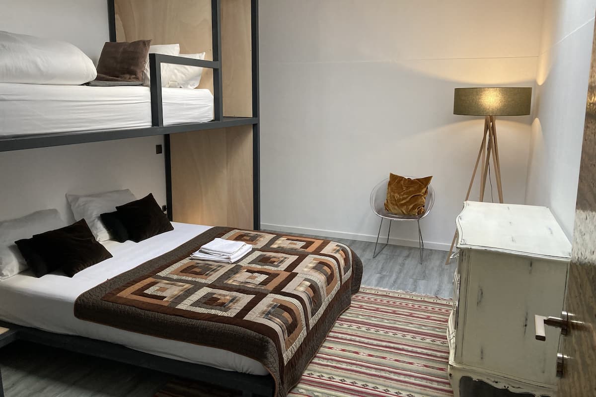 Quarto de hotel pequeno com beliche, uma cama de solteiro, tapete estampado, uma cadeira e um abajur.