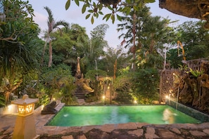 Uma área de piscina ao ar livre tranquila, rodeada por vegetação exuberante, com escadas de pedra que levam a um gazebo e um lampião de pedra.