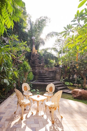 Um pátio com cadeiras brancas e detalhadas, uma mesa, vegetação exuberante e uma escadaria de pedra que leva a uma estrutura semelhante a um templo.