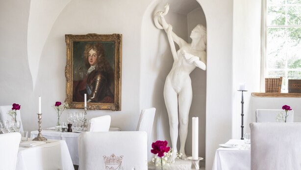 Uma área de refeições com toalhas de mesa brancas, castiçais e uma estátua de mulher.