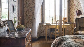 Um quarto com papel de parede floral, uma escrivaninha de madeira, uma cadeira, um abajur e uma cama coberta de pele.