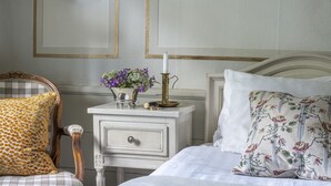 Quarto bem arrumado com cama, mesa de cabeceira com um vaso de flores, cadeira com almofada estampada e um candelabro decorativo.