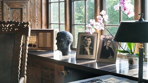 Uma escrivaninha de madeira com um busto, fotografias emolduradas e um vaso de planta perto de uma janela com vista para o verde.
