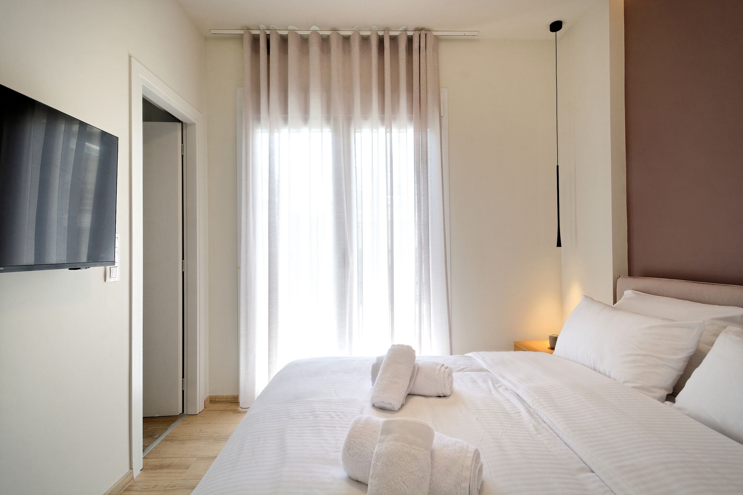 Quarto de hotel com cama, televisão presa na parede, janela com cortinas e mesa de cabeceira com abajur.