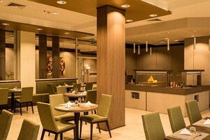 Um restaurante moderno com mesas redondas, cadeiras estofadas em verde e uma coluna central de madeira.