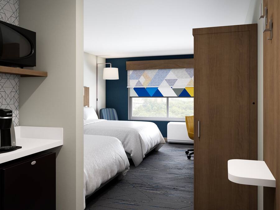 Quarto de hotel com cama, escrivaninha, cadeira, televisão e janela com cortina geométrica.