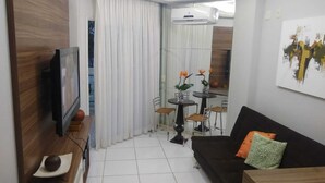 Sala de estar com sofá preto, pequena mesa de jantar com cadeiras, televisão à esquerda e cortina branca separando a área de estar da área de jantar.