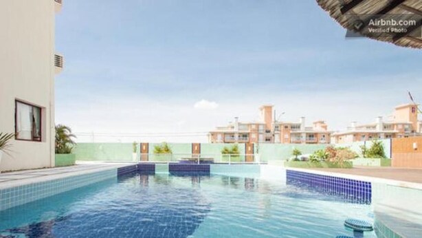 Piscina com água azul cristalina, rodeada por um deck revestido em azulejos e um muro baixo.