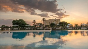 Piscina com espreguiçadeiras e guarda-sóis, cercada por árvores e um edifício com janelas em arco.