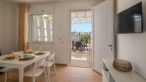 Uma área de refeições com uma mesa posta para quatro pessoas, uma televisão na parede e vista para um pátio externo.