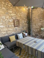 Área de estar ao ar livre com paredes de pedra, um sofá e uma mesa, um quadro na parede e uma pequena mesa com uma garrafa e flores.