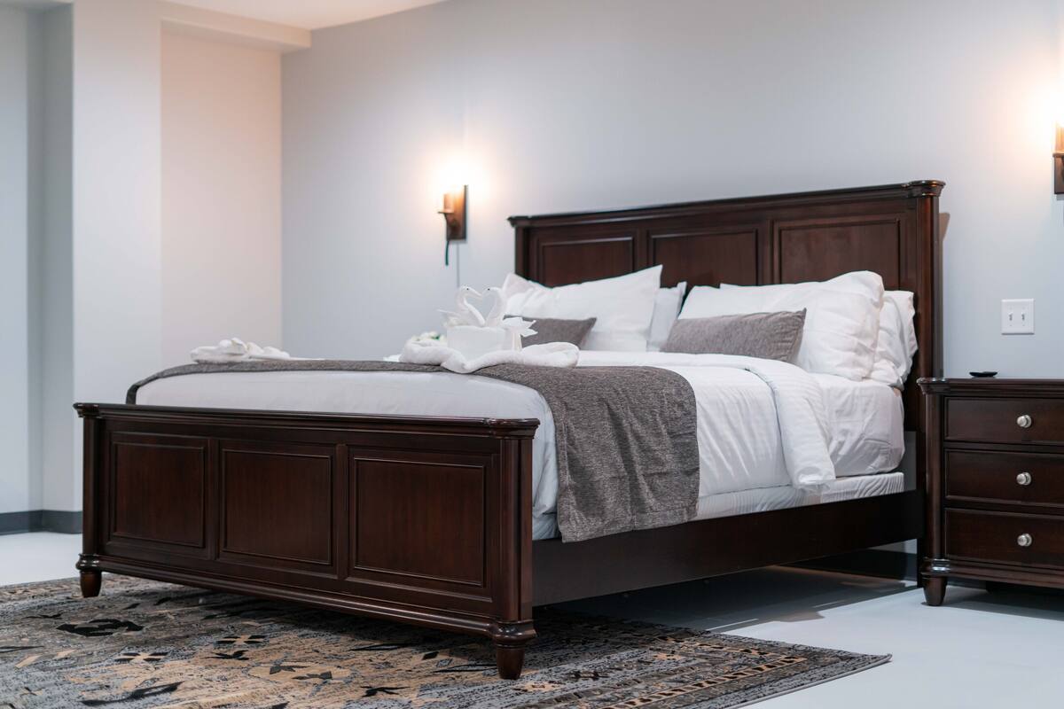 Quarto com cama de madeira escura, roupa de cama branca e tapete estampado.