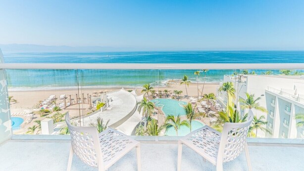 Varanda com vista para um resort na praia, incluindo área de piscina e palmeiras.