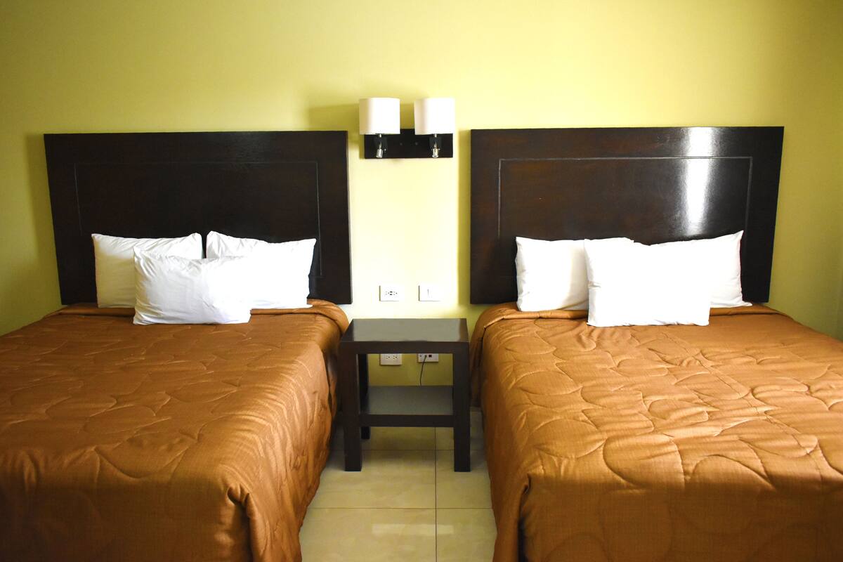 Duas camas de solteiro com roupa de cama castanha, uma mesa de cabeceira com abajur entre elas, e uma luminária fixada na parede.