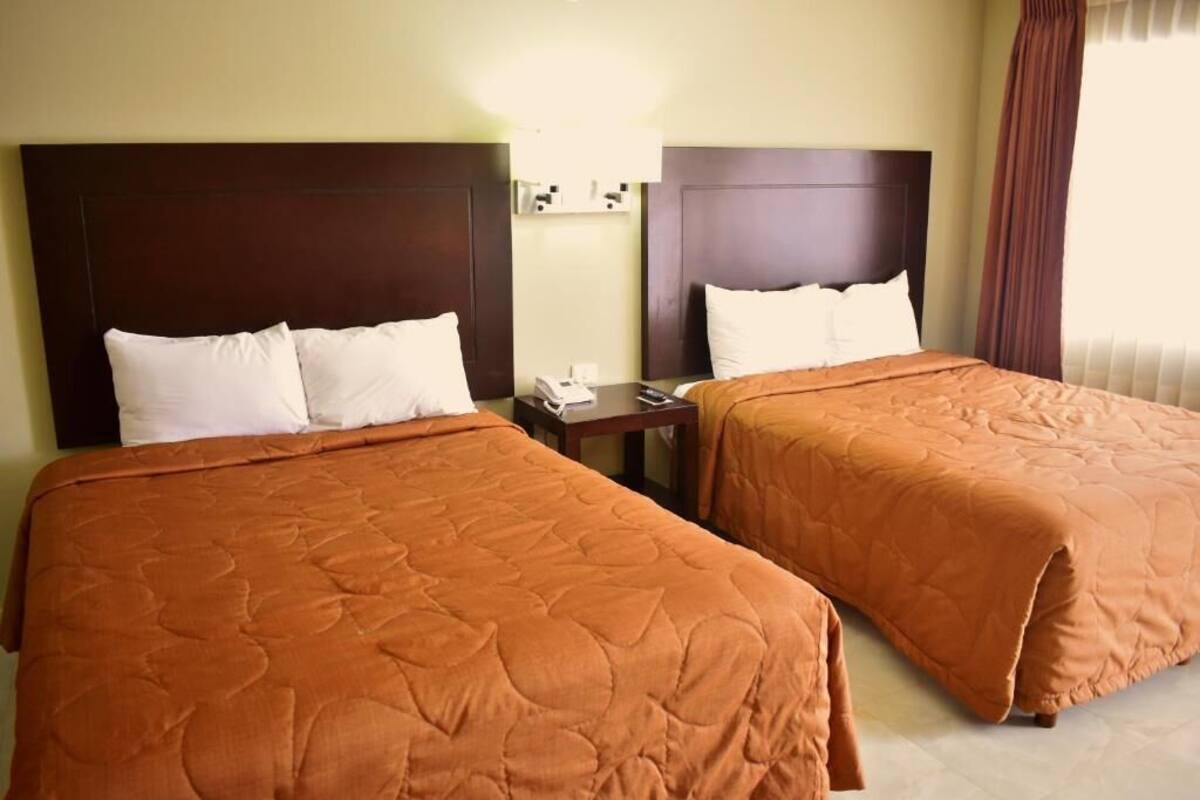Quarto de hotel com duas camas, cada uma com colcha acolchoada marrom e almofadas brancas. Um criado-mudo entre as camas possui um telefone e um abajur.