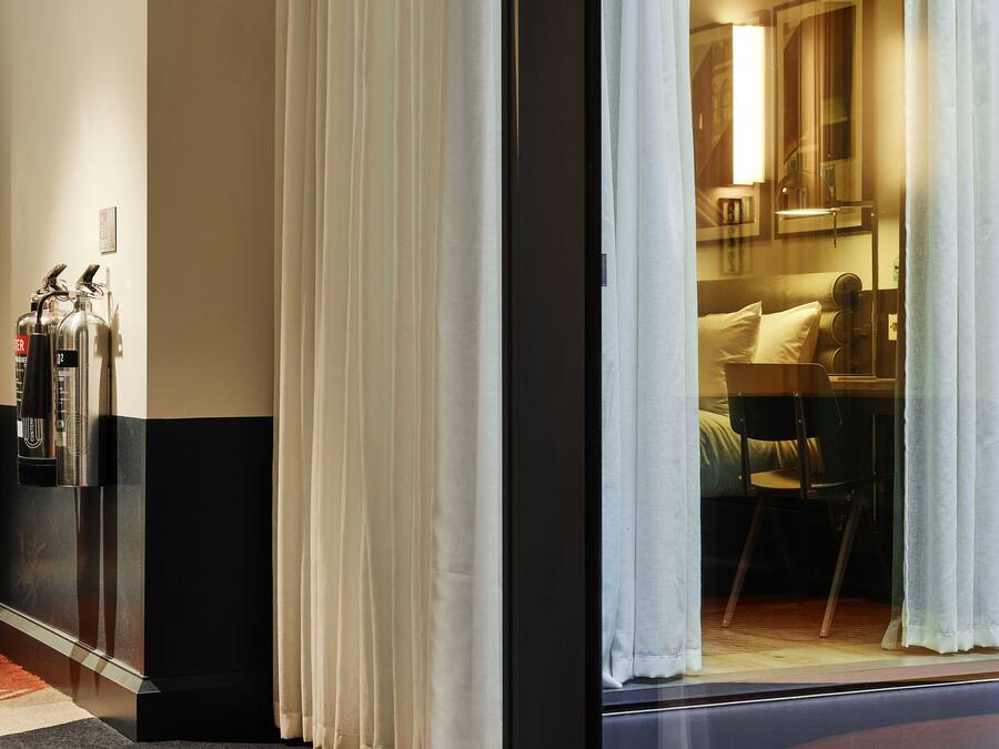 Quarto de hotel com uma grande porta de correr de vidro que dá para uma varanda, uma cama com travesseiro, uma cadeira e uma escrivaninha com abajur. Há dois extintores montados na parede.