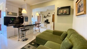 Cozinha moderna com bancada preta, eletrodomésticos de aço inoxidável e área de bar. Há um sofá verde na sala de estar e uma moldura com um quadro na parede.