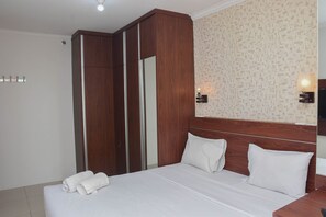 Uma cama bem arrumada com lençóis e almofadas brancos, um guarda-roupa de madeira e um aparelho de luz fixado na parede.