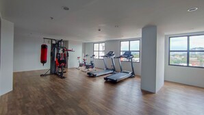 Uma academia ampla com piso de madeira, diversos equipamentos de exercício e janelas grandes com vista para a cidade.