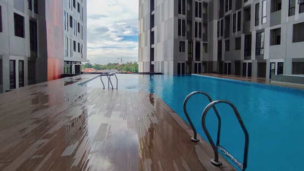 Piscina com deck de madeira e grades de metal, rodeada por edifícios modernos com grandes janelas.