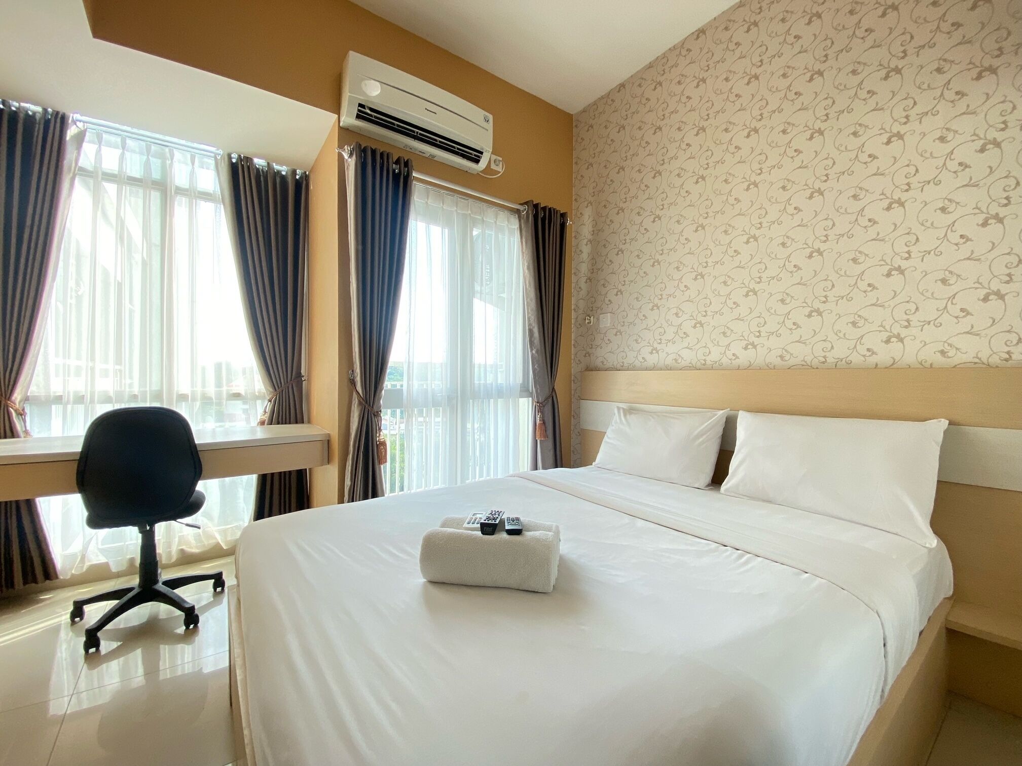 Quarto de hotel com cama, escrivaninha, aparelho de ar condicionado e janela com cortinas.