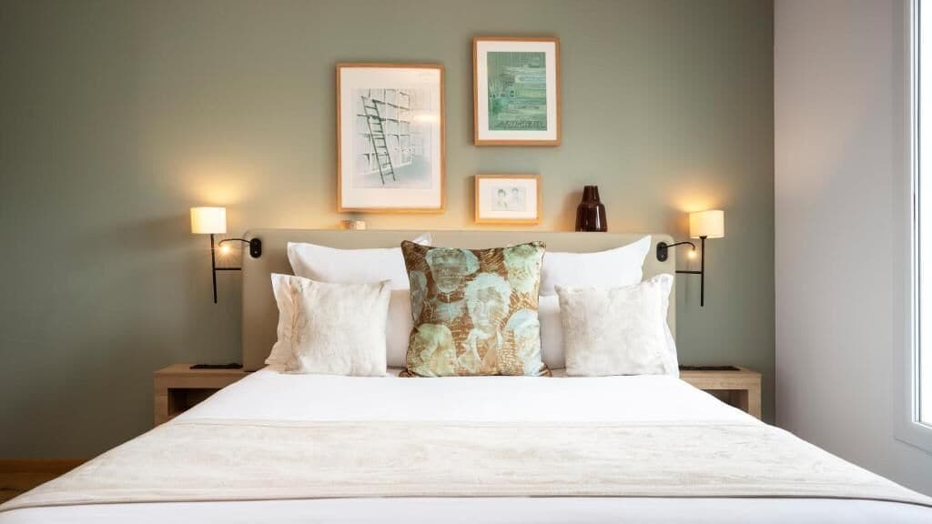 Cama com roupa de cama branca, duas mesas de cabeceira, luminárias fixadas na parede e quadros na parede.
