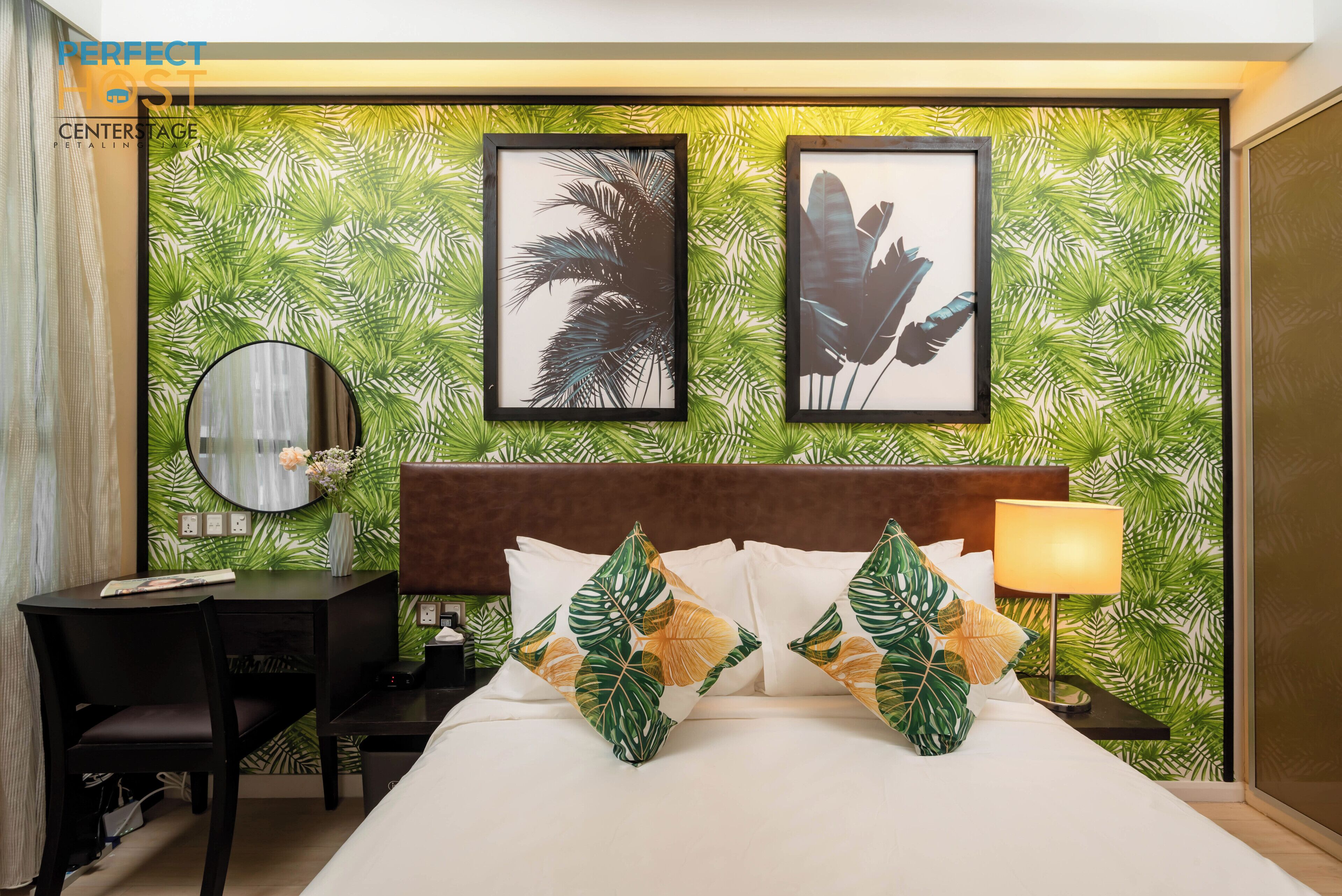 Quarto de hotel com cama, escrivaninha, cadeira, mesa de cabeceira, abajur e espelho. O papel de parede apresenta um padrão de folhagem tropical.