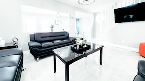 Uma sala de estar moderna com um sofá de couro preto, uma mesa de centro de vidro e uma televisão de tela plana fixada na parede.