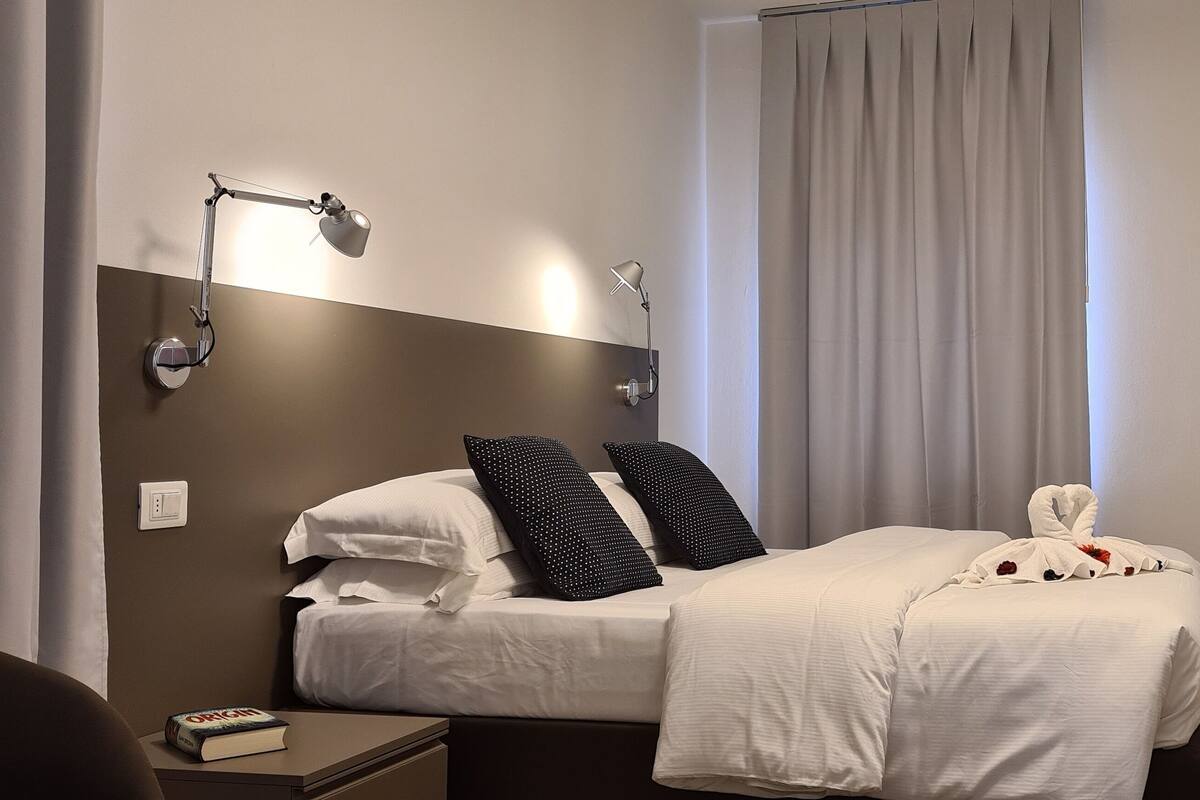 Cama bem arrumada com roupa de cama branca e duas almofadas pretas, uma mesa de cabeceira com um livro e uma luminária fixada na parede.