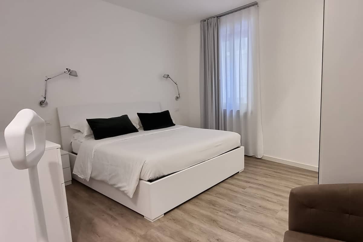 Quarto moderno com cama, duas luminárias de cabeceira, uma cadeira e uma janela com cortinas.