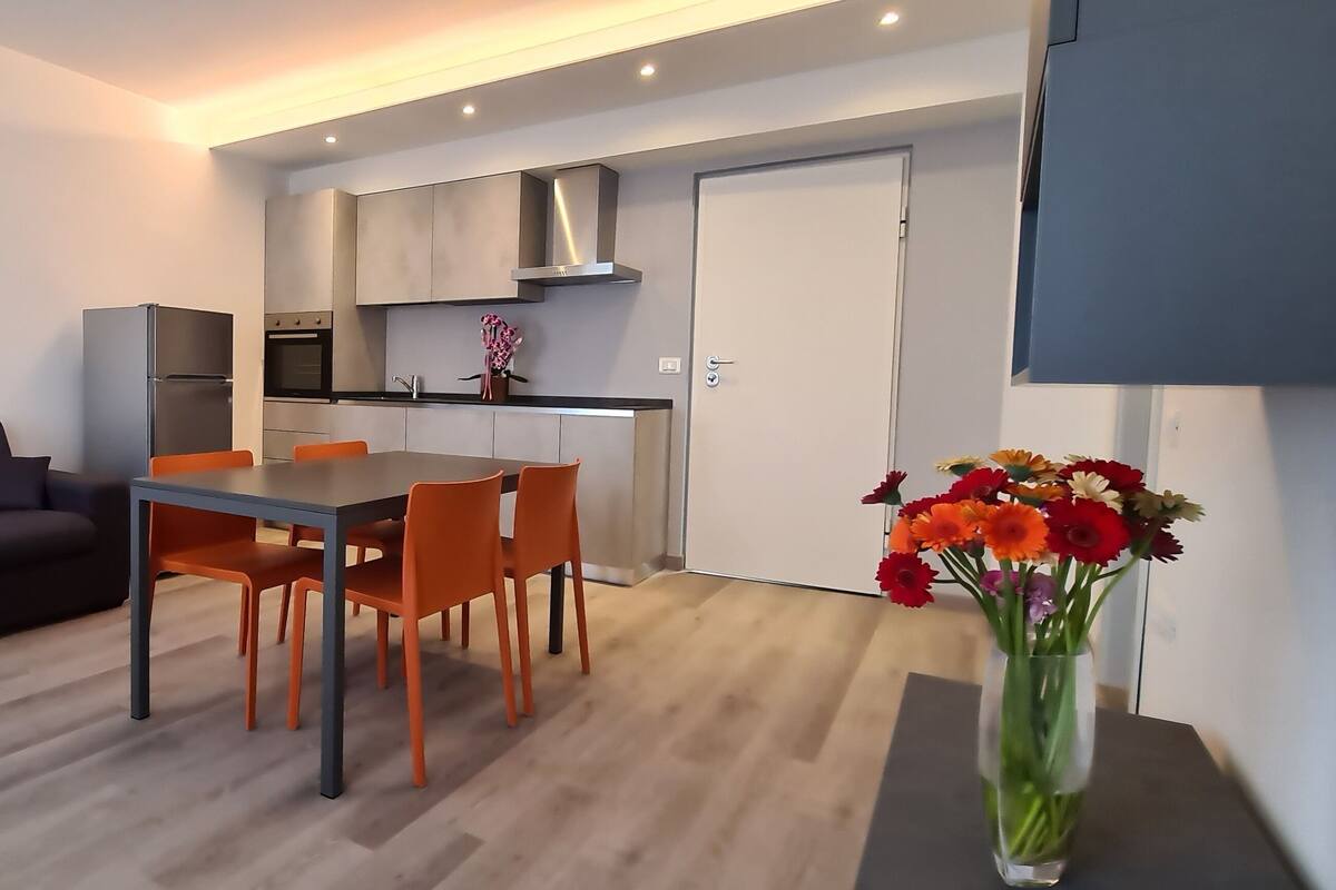 Uma sala de estar moderna com sofá, mesa de jantar e cadeiras, uma cozinha equipada e um vaso de flores.