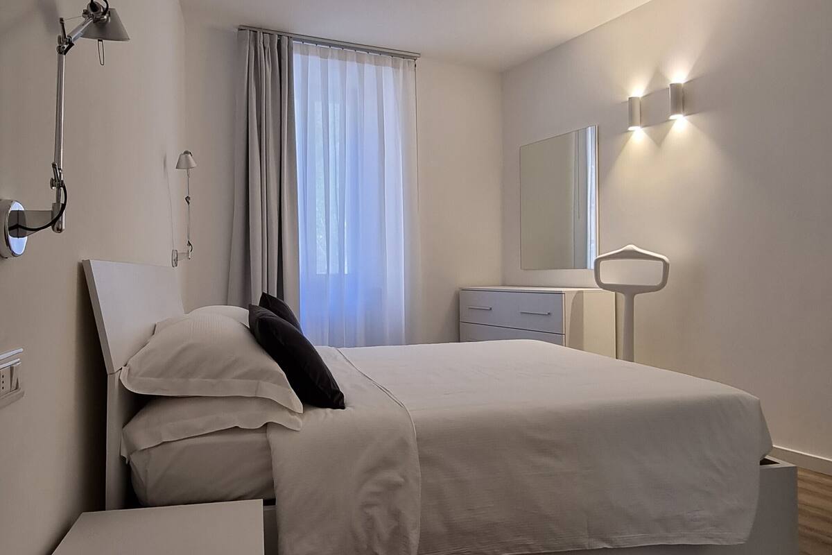 Quarto bem arrumado com cama, mesa de cabeceira, abajur e um espelho na parede.