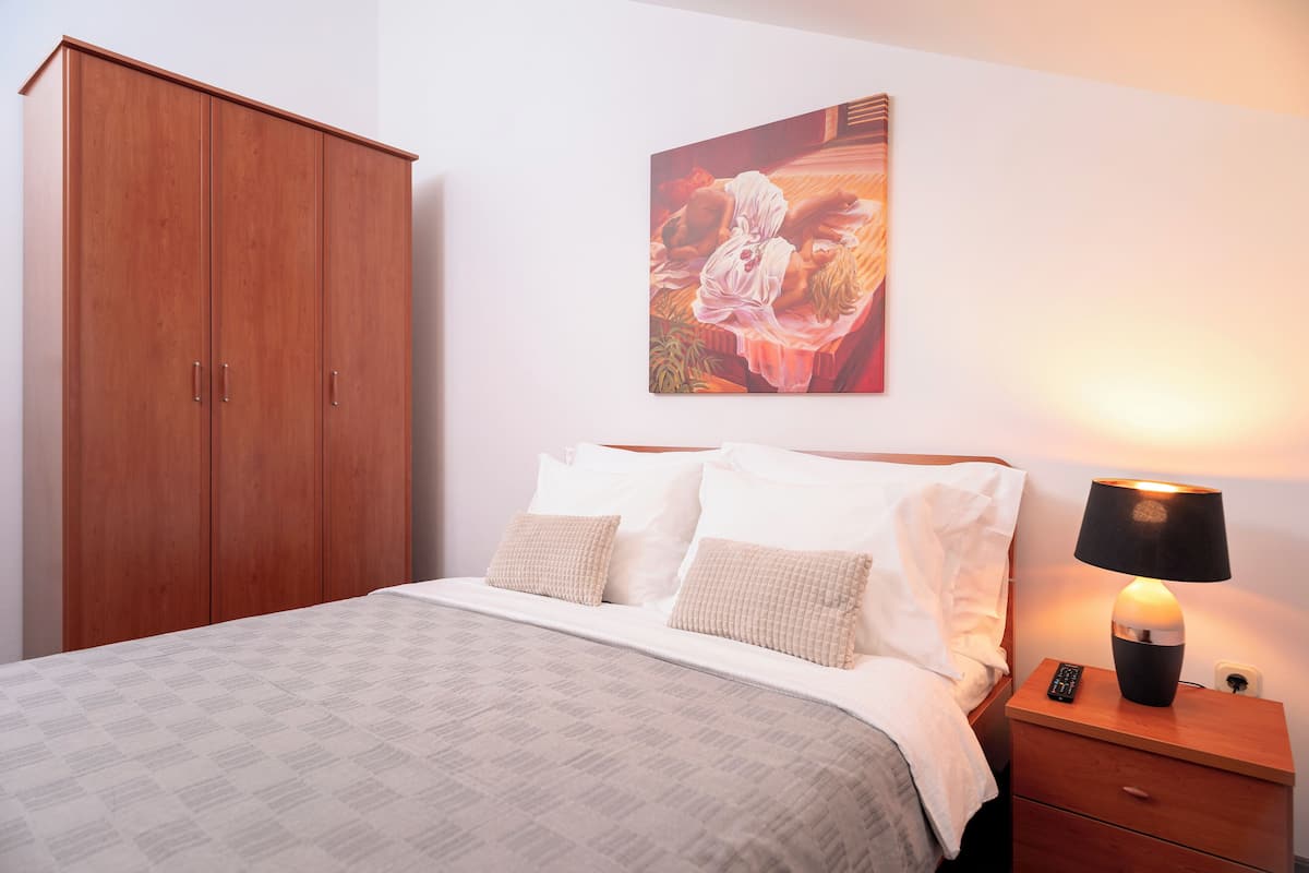Quarto com cama, mesa de cabeceira, luminária, pintura e guarda-roupa.