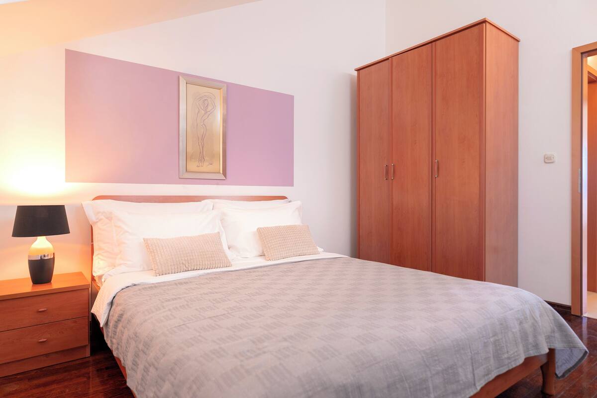 Uma cama bem arrumada com edredom branco e duas almofadas, mesa de cabeceira de madeira com abajur, guarda-roupa espaçoso e uma obra de arte emoldurada na parede.